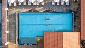 Vista aérea de uma piscina retangular com duas pessoas nadando, cercada por um deque de madeira e uma parede de tijolos.