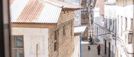 Uma rua estreita com edifícios em ambos os lados, uma varanda com grades e uma pessoa caminhando pela rua.