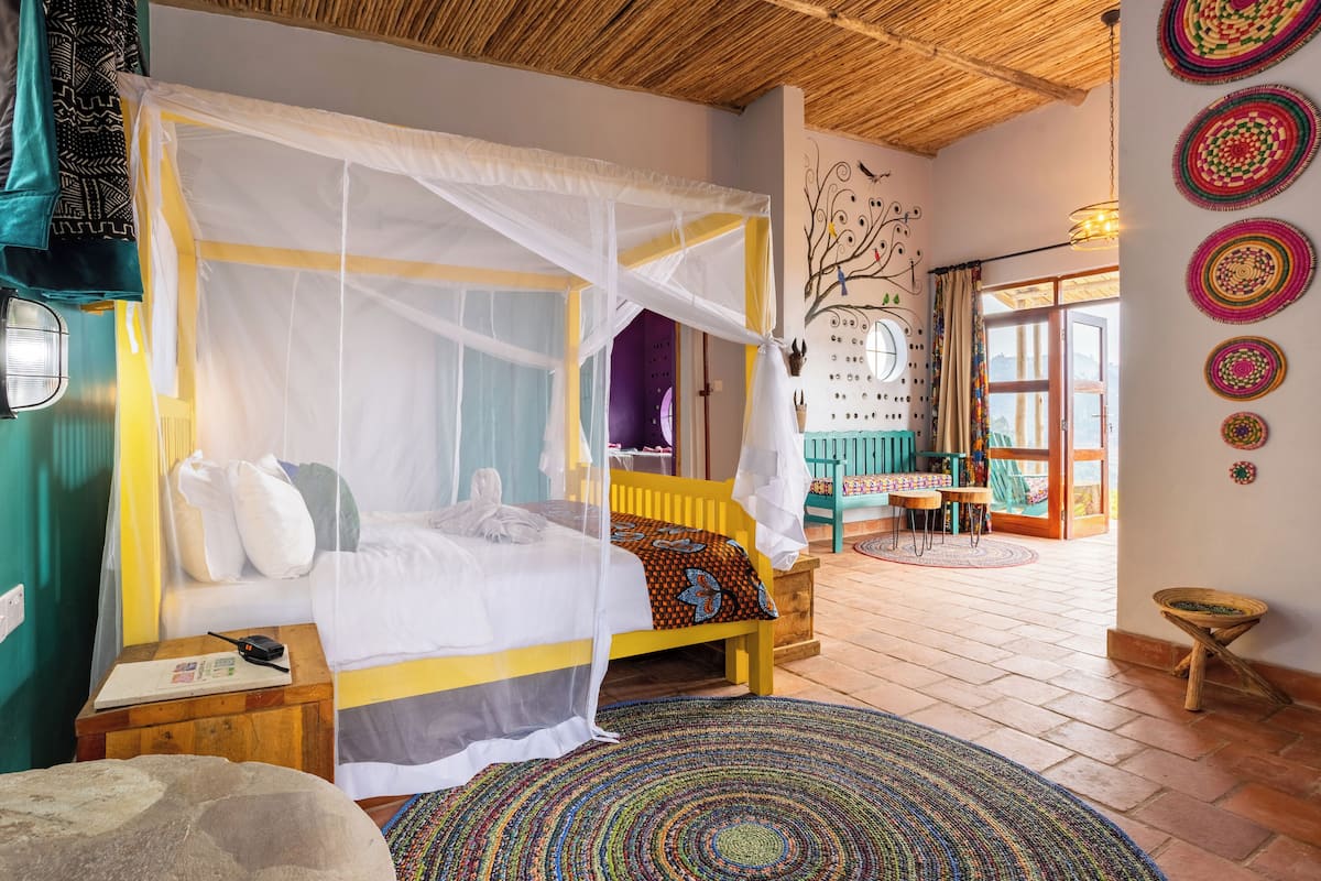Quarto com cama sob uma mosquiteira, mesa de cabeceira de madeira, tapete colorido e vista para o exterior.