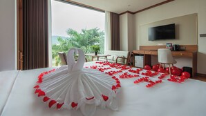 Quarto de hotel com cama decorada com toalha em formato de cisne e pétalas de rosa vermelha, uma mesa com computador, uma cadeira e uma televisão na parede.