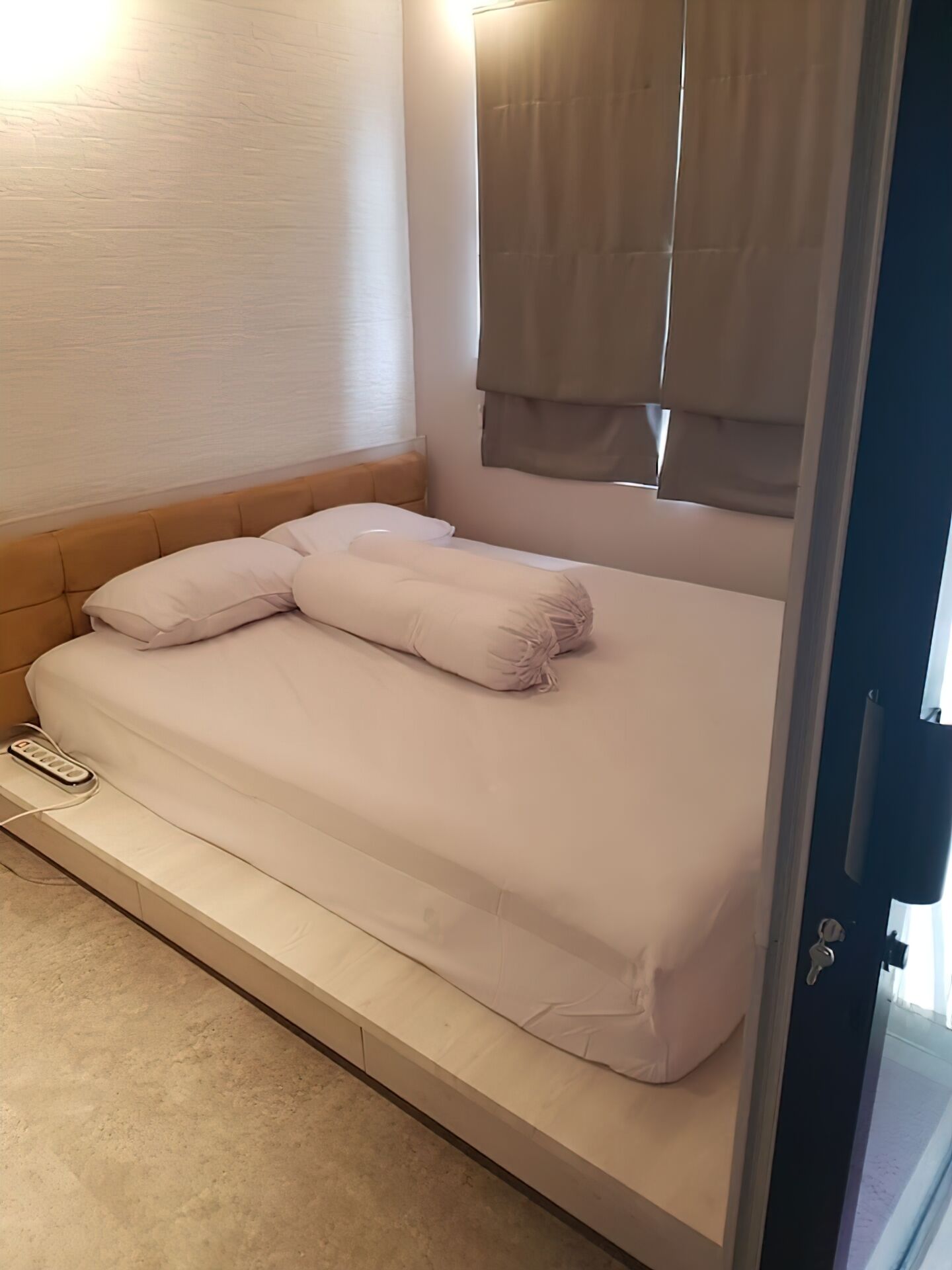 Uma cama bem arrumada com cabeceira, mesa de cabeceira com tomada e janela com cortinas.