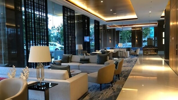 Um hall de hotel moderno, com um balcão de recepção extenso, área de estar com sofás e cadeiras, e janelas amplas com vista para a rua.