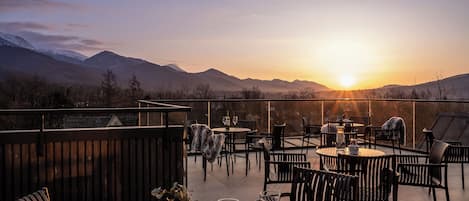 Área de refeições ao ar livre com mesas e cadeiras, com vista para uma paisagem montanhosa durante o pôr do sol.