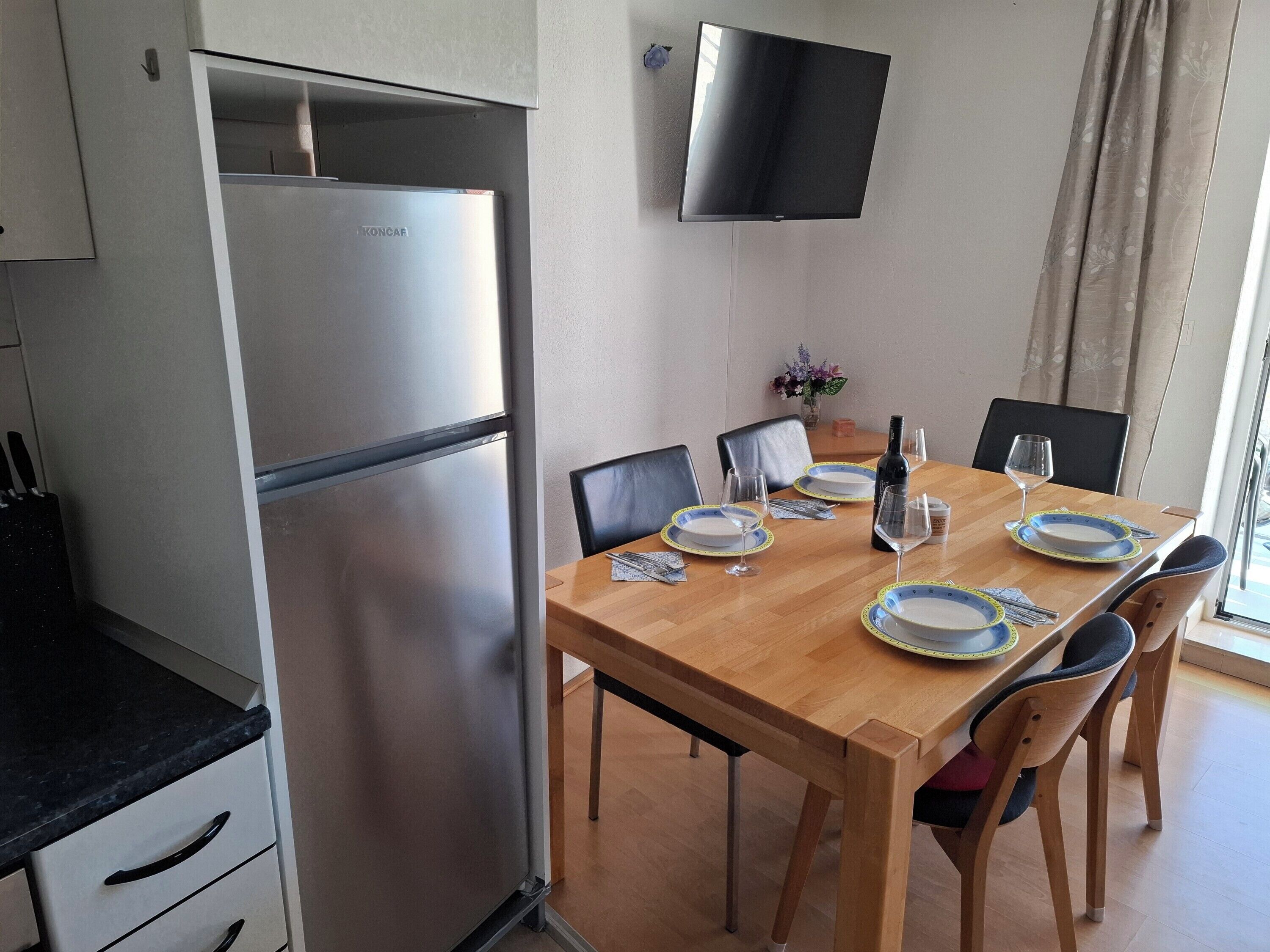 Uma sala de jantar com mesa de madeira posta para quatro pessoas, uma televisão de tela plana na parede e uma geladeira de aço inoxidável.