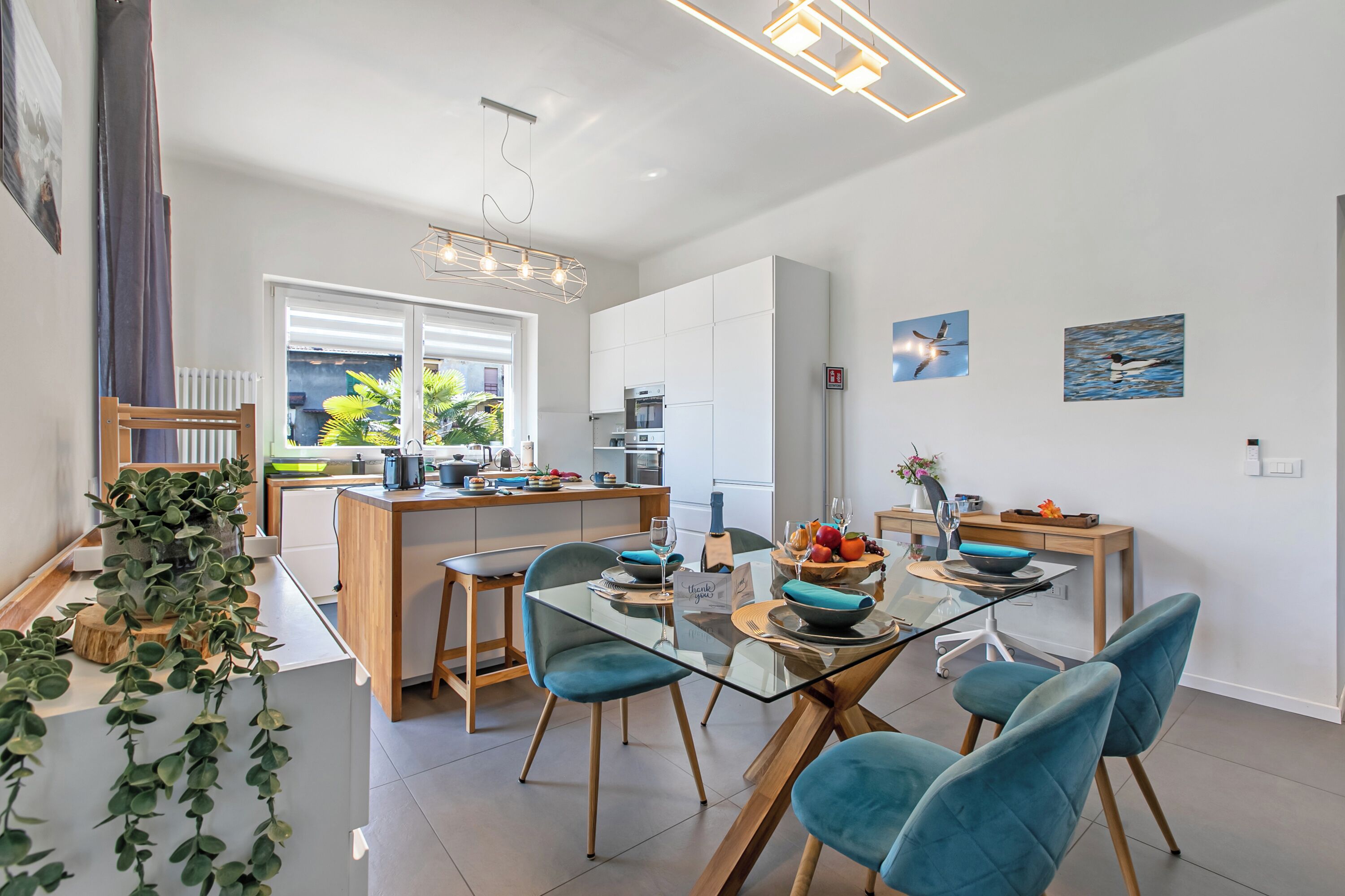 Cozinha moderna com armários brancos, área de refeições com mesas de vidro e cadeiras verde-água, e vista para o exterior através de grandes janelas.
