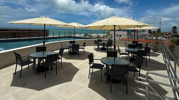 Uma área de piscina no terraço com mesas, cadeiras, guarda-sóis e vista para o oceano.