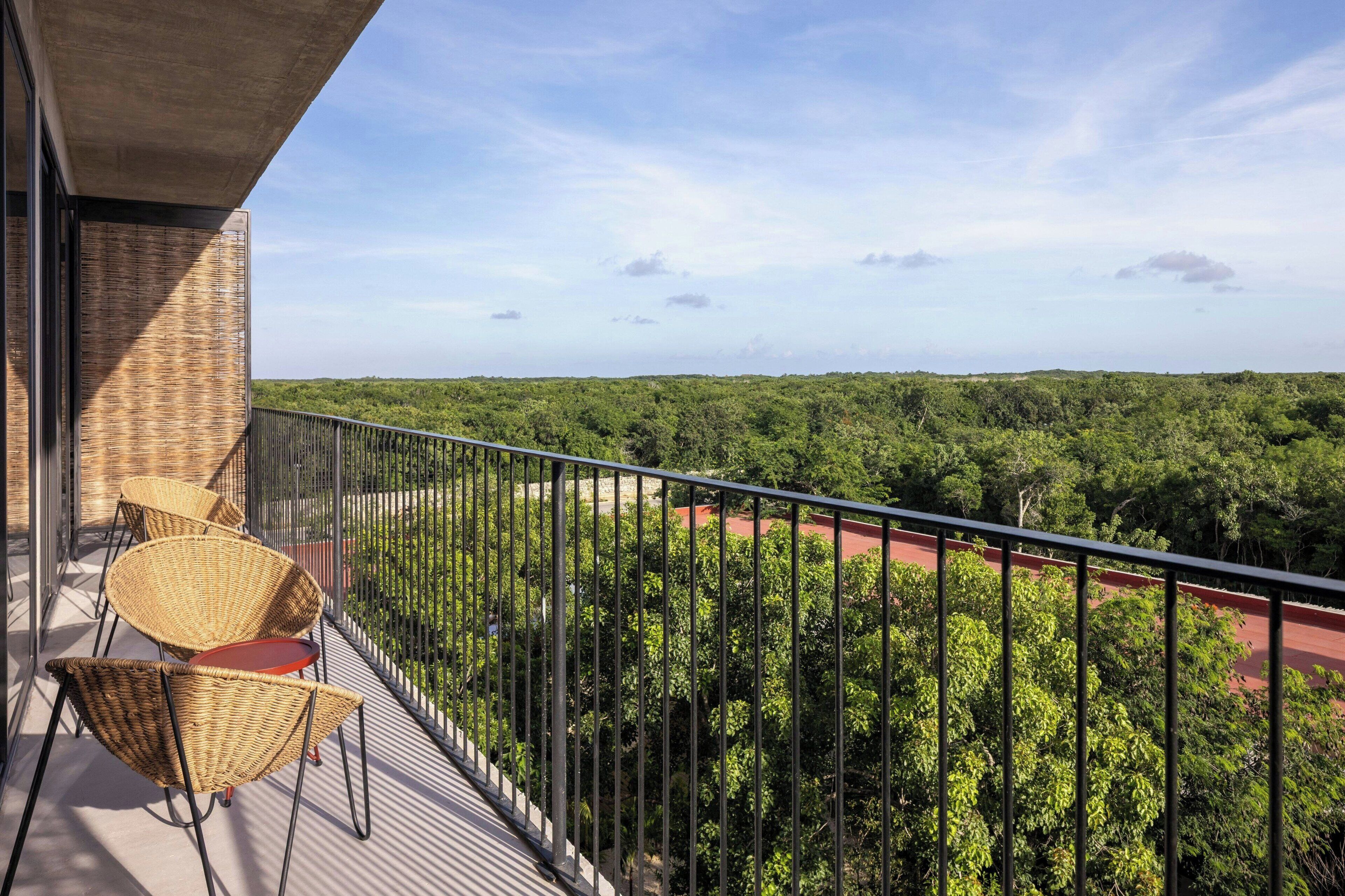 Varanda com cadeiras de vime e uma mesa vermelha, com vista para uma floresta densa e uma estrada vermelha.