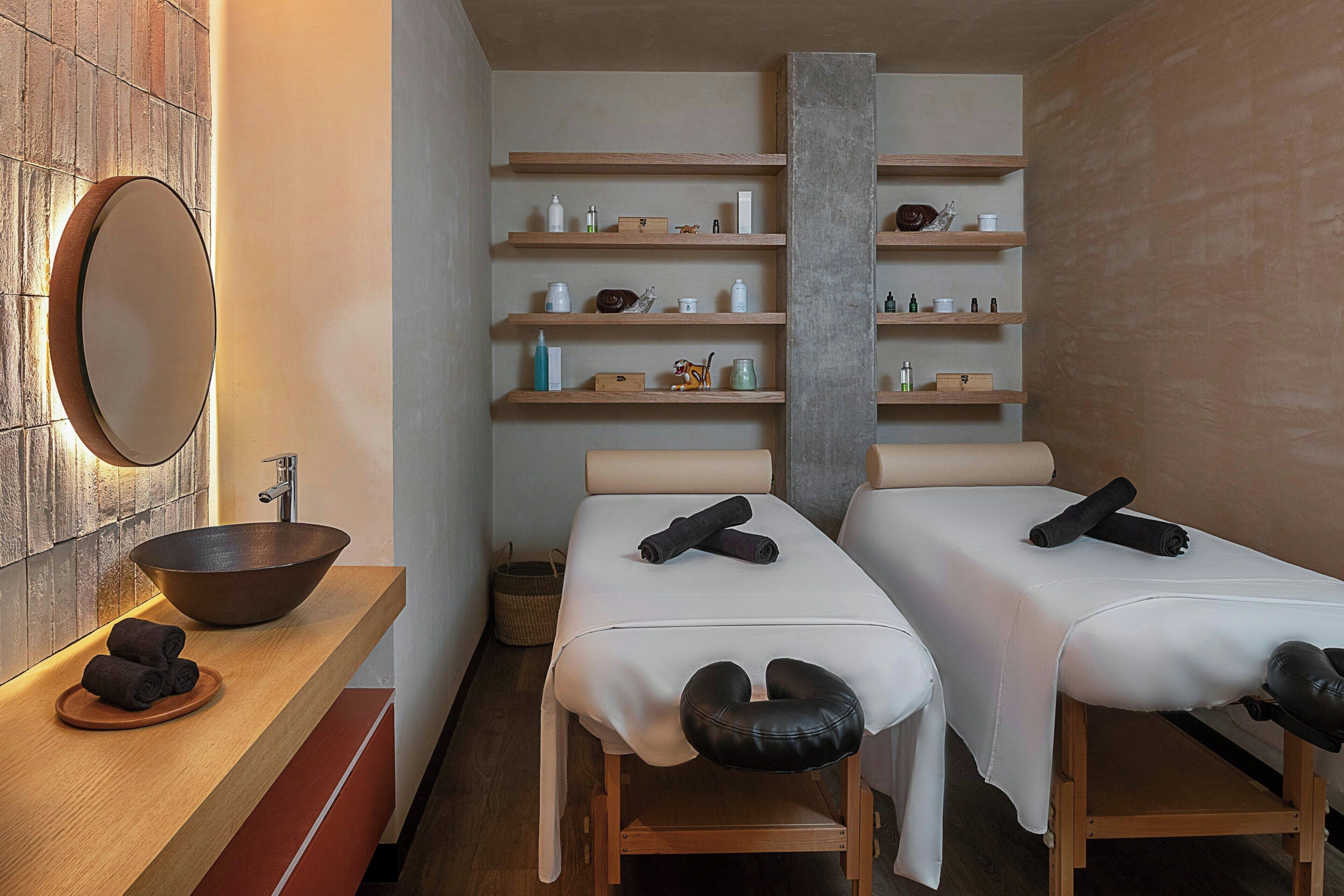 Sala de spa com duas mesas de massagem, uma prateleira de madeira com frascos, um espelho grande e uma tigela de madeira no balcão.
