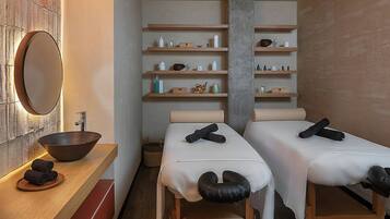 Sala de spa com duas mesas de massagem, uma prateleira de madeira com frascos, um espelho grande e uma tigela de madeira no balcão.