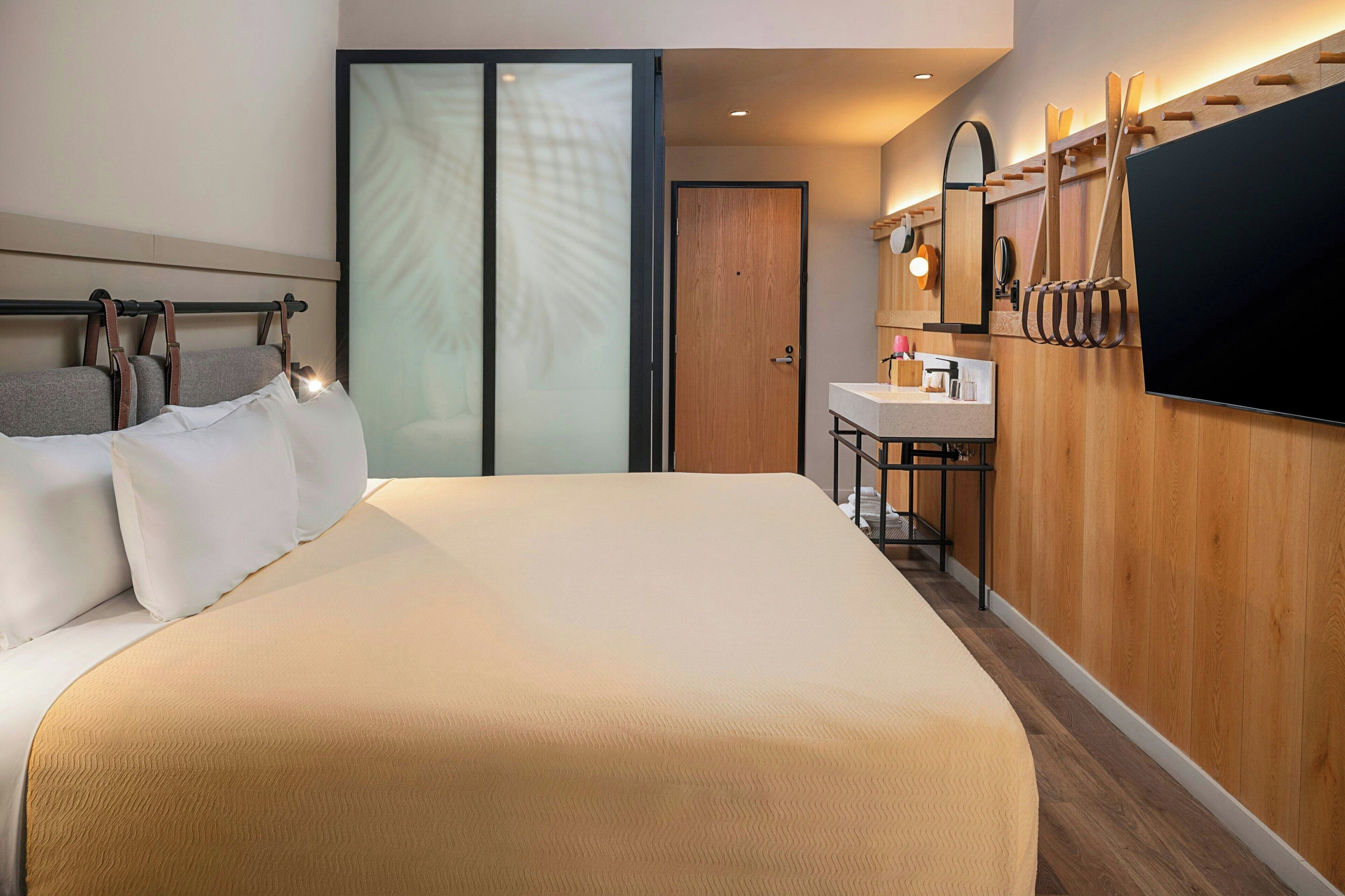 Quarto de hotel com uma cama, parede de madeira, espelho, televisão e um pequeno banheiro.