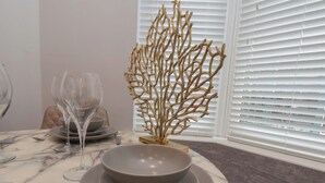 Uma mesa posta com uma tigela, pratos e talheres, um ramo dourado decorativo e uma janela com persianas.