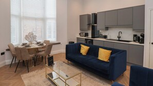 Uma sala de estar moderna com uma mesa de centro de vidro, um sofá azul com almofadas amarelas, e uma área de jantar com uma mesa e cadeiras brancas.