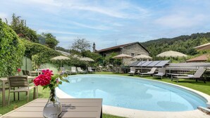 Piscina com espreguiçadeiras e guarda-sóis, cercada por vegetação e edifícios de pedra.