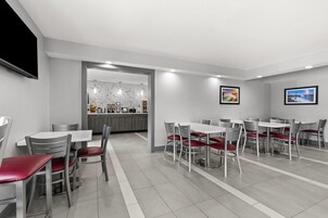 Uma sala de jantar moderna com várias mesas e cadeiras, uma cozinha ao fundo e quadros nas paredes.