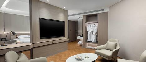 Quarto de hotel moderno com TV de tela plana, uma escrivaninha com abajur, uma mesa de centro redonda, duas poltronas, um closet espaçoso com roupões pendurados e piso de madeira.