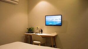 Quarto de hotel com cama, escrivaninha, cadeira, televisão fixada na parede e um vaso de planta.