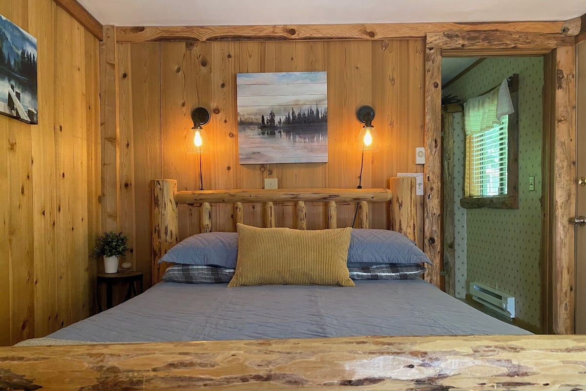 Quarto com painéis de madeira, uma cama, duas luminárias fixadas na parede e um quadro sobre a cama.