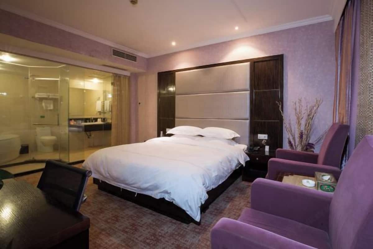 Quarto de hotel com cama, escrivaninha, cadeira, box de chuveiro de vidro e um espelho grande.