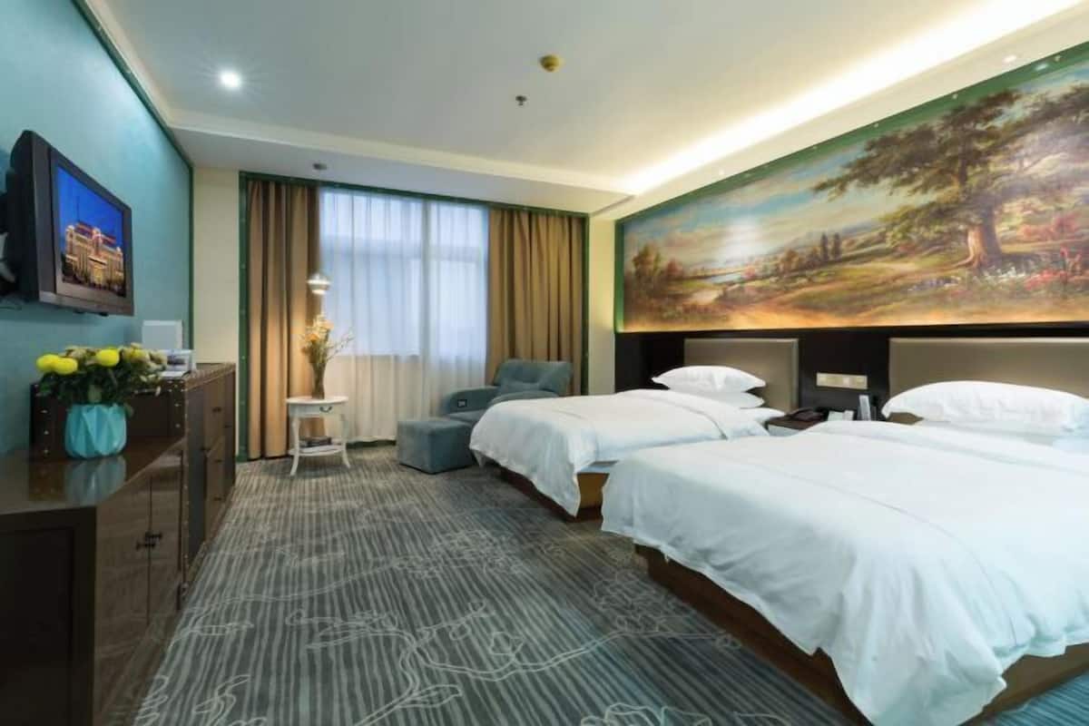 Quarto de hotel com duas camas, uma escrivaninha, uma cadeira, um quadro na parede e uma janela com cortinas.