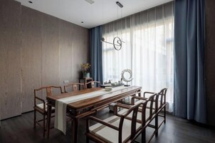 Uma sala de jantar com mesa e cadeiras de madeira, um vaso com flores e vista através de cortinas leves.