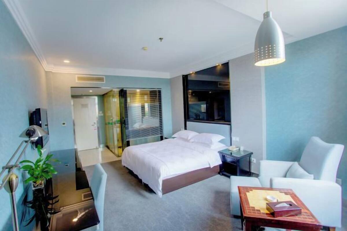 Quarto de hotel com cama, escrivaninha, cadeira, planta e luminária suspensa.