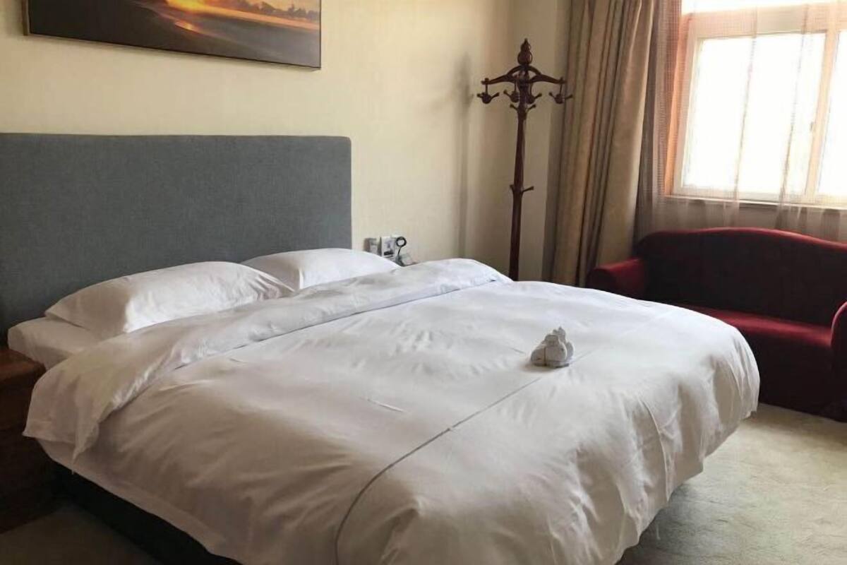 Uma cama bem arrumada com lençóis brancos, uma cadeira vermelha e um cabideiro.