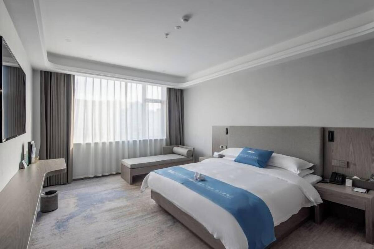 Quarto de hotel moderno com uma cama grande, uma mesinha de cabeceira, uma escrivaninha e uma janela ampla com cortinas.