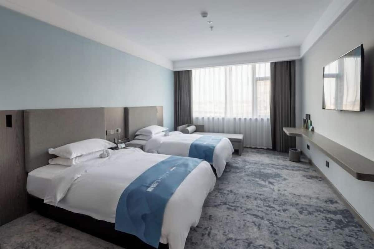 Quarto de hotel com duas camas, uma janela grande com cortinas, uma TV de tela plana e uma mesinha de cabeceira.