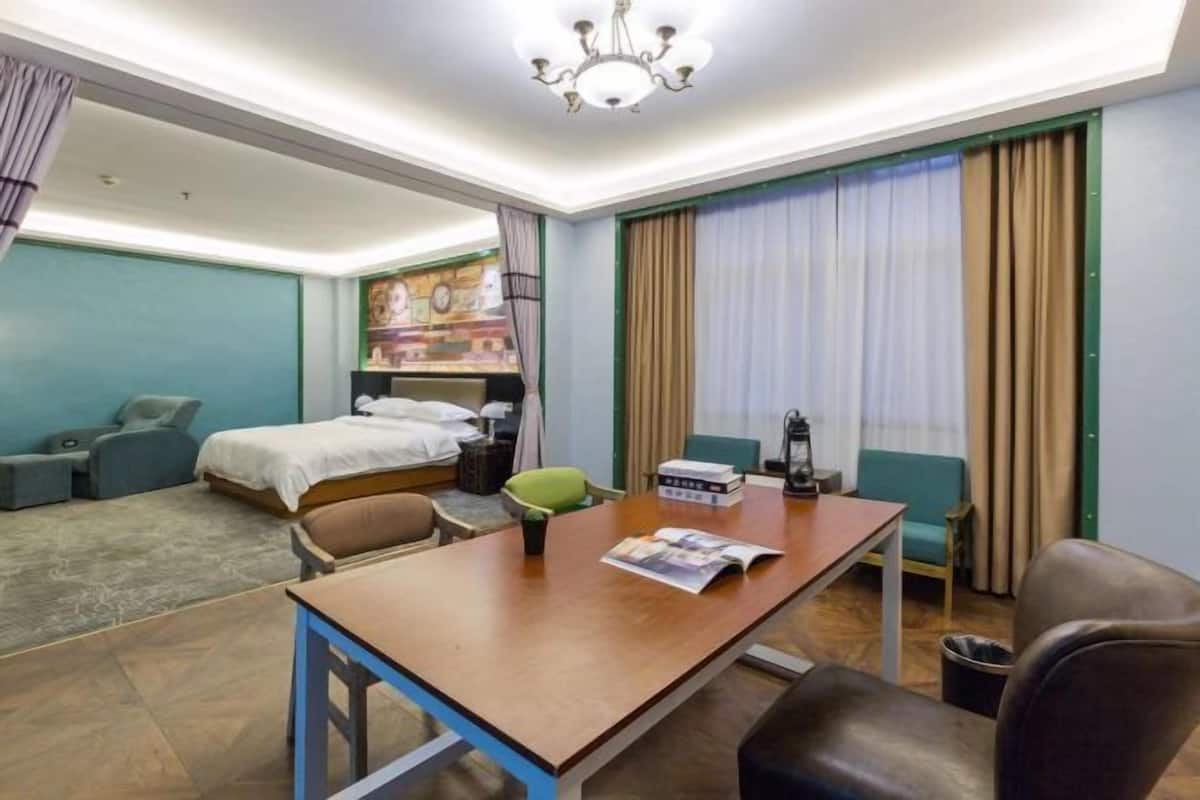 Quarto de hotel com cama, escrivaninha, cadeira, mesa pequena, luminária, estante de livros e janela com cortinas.