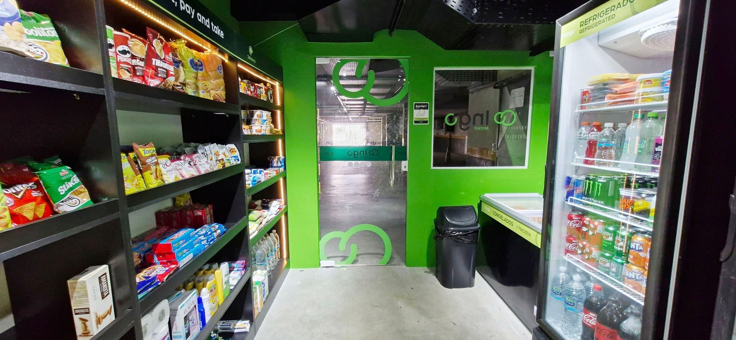 Corredor estreito com prateleiras cheias de lanches e bebidas, uma lixeira e uma área refrigerada para bebidas.