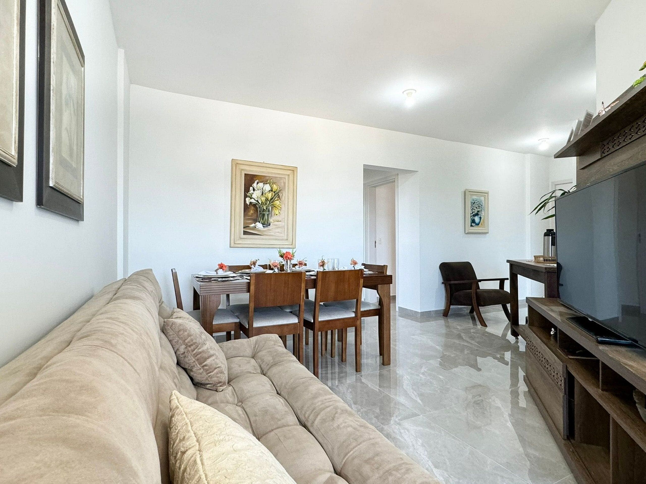 Uma sala de estar com um sofá bege, uma mesa de jantar com cadeiras, uma televisão e quadros nas paredes.