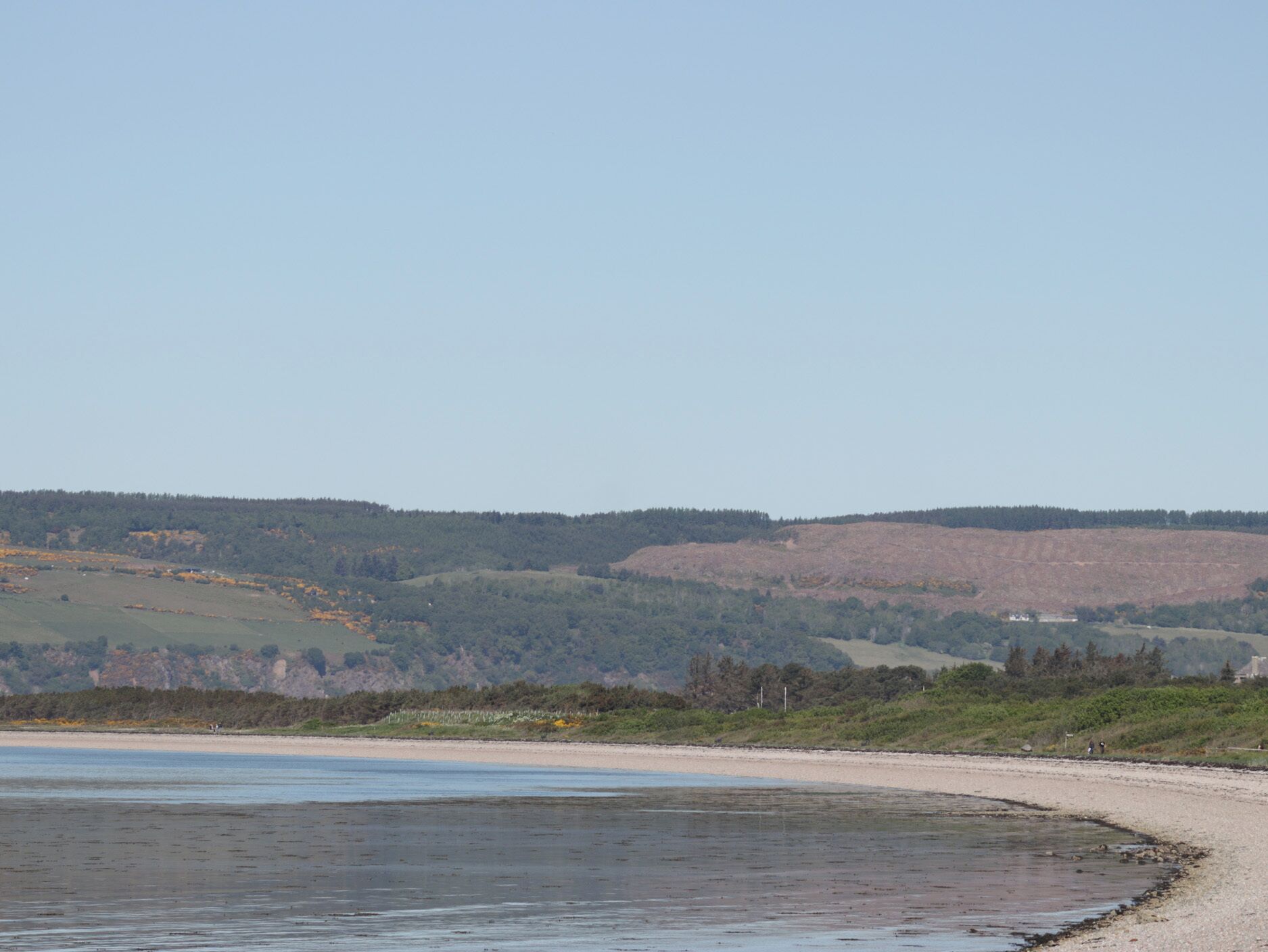 Paisagem costeira com uma praia arenosa, mar calmo e colinas ondulantes ao fundo.