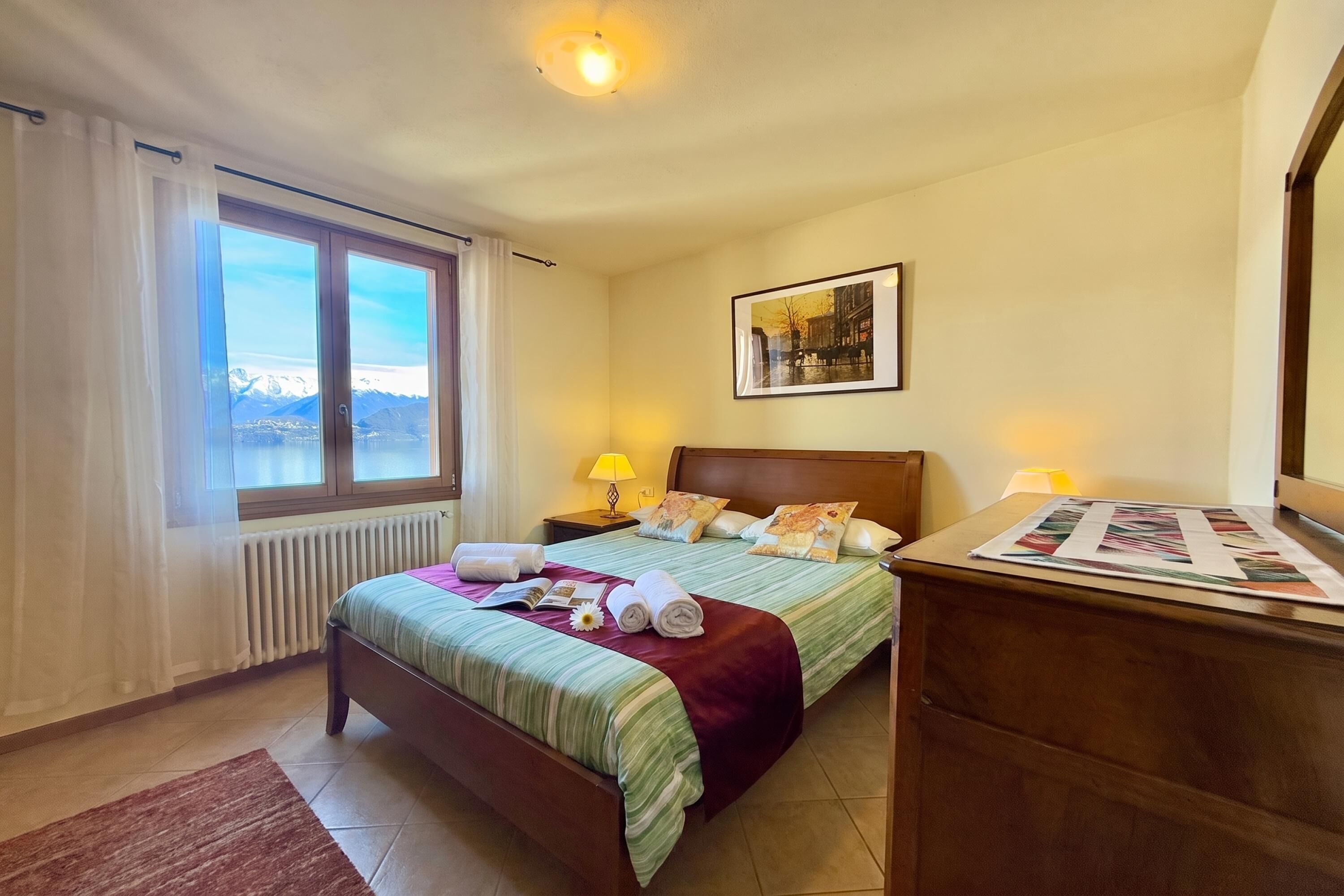 Quarto de hotel com uma cama, mesa de cabeceira com abajur, janela com cortinas, radiador e um quadro na parede.