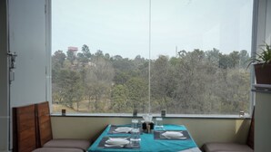 Uma área de jantar com uma mesa posta para dois, uma janela com vista para árvores e um edifício distante, e uma planta em vaso à direita.
