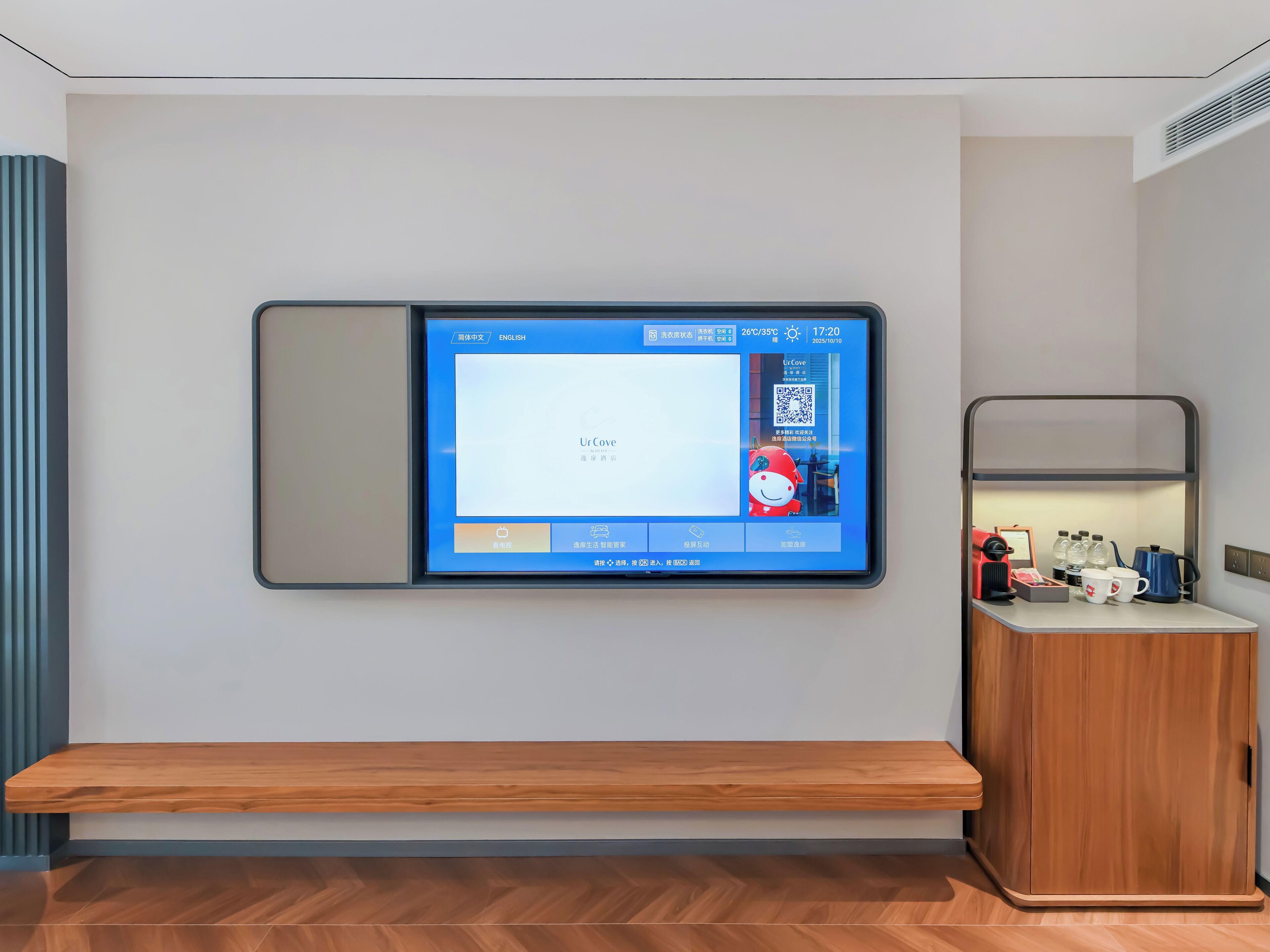 Quarto moderno com tela digital fixada exibindo um código QR, um banco de madeira e uma pequena mesa com chaleira e xícaras.