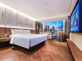 Quarto de hotel moderno com uma cama grande, uma escrivaninha, uma cadeira, uma televisão e vista para a cidade.