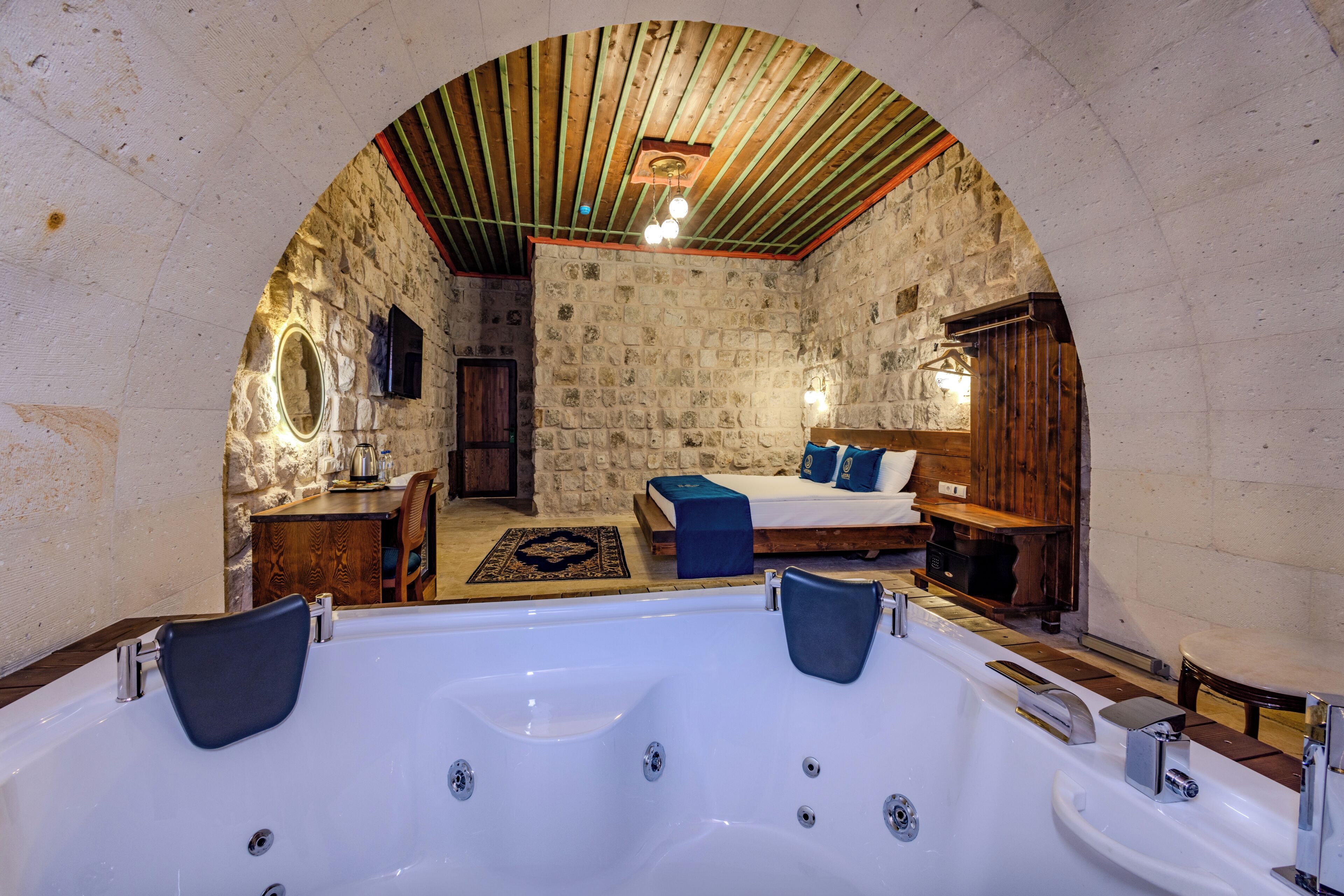 Quarto com paredes de pedra, teto de madeira, uma banheira grande, uma cama com roupa de cama azul e um armário de madeira.