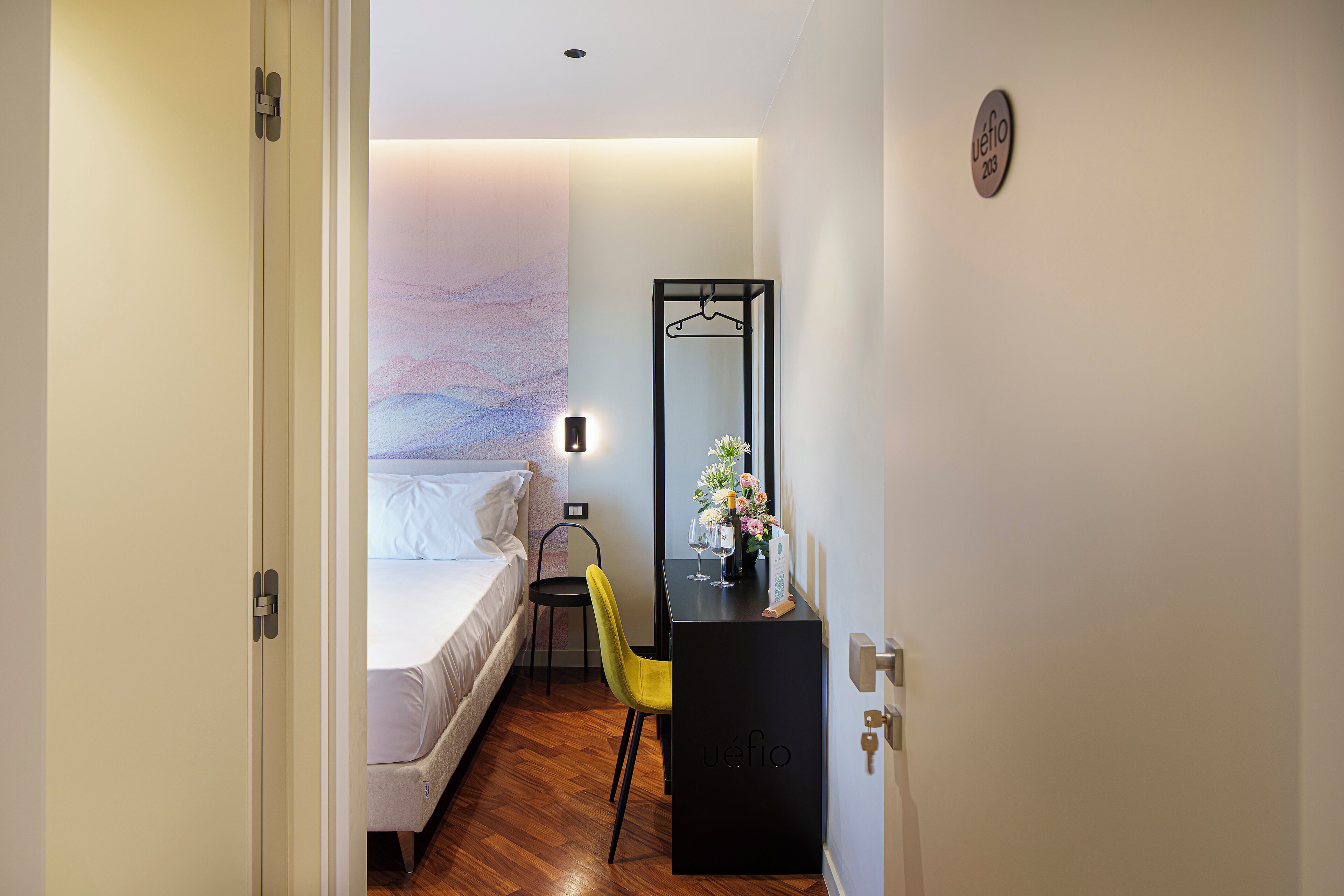 Quarto de hotel com cama, escrivaninha, cadeira, espelho e um objeto decorativo na parede.