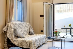 Uma sala de estar moderna com um sofá estampado, uma mesinha redonda com uma planta em vaso e uma porta de vidro deslizante que dá para uma área externa com mesa e cadeiras.
