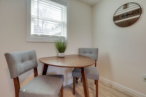 Uma sala de jantar com uma mesa de madeira redonda, duas cadeiras estofadas, um vaso com planta e um relógio de parede.