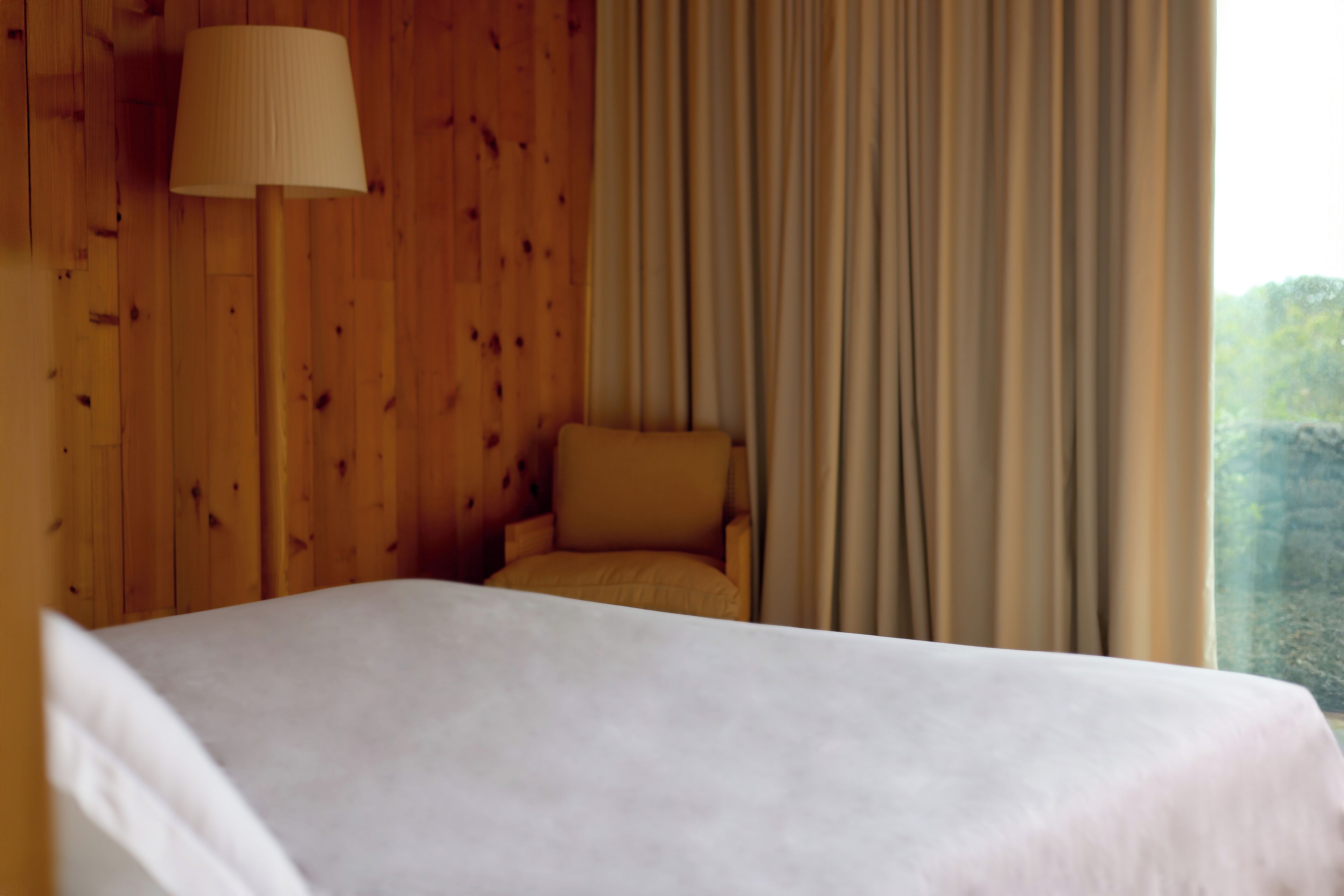 Quarto de hotel com cama, poltrona, luminária, paredes de madeira e vista pela janela com cortinas.