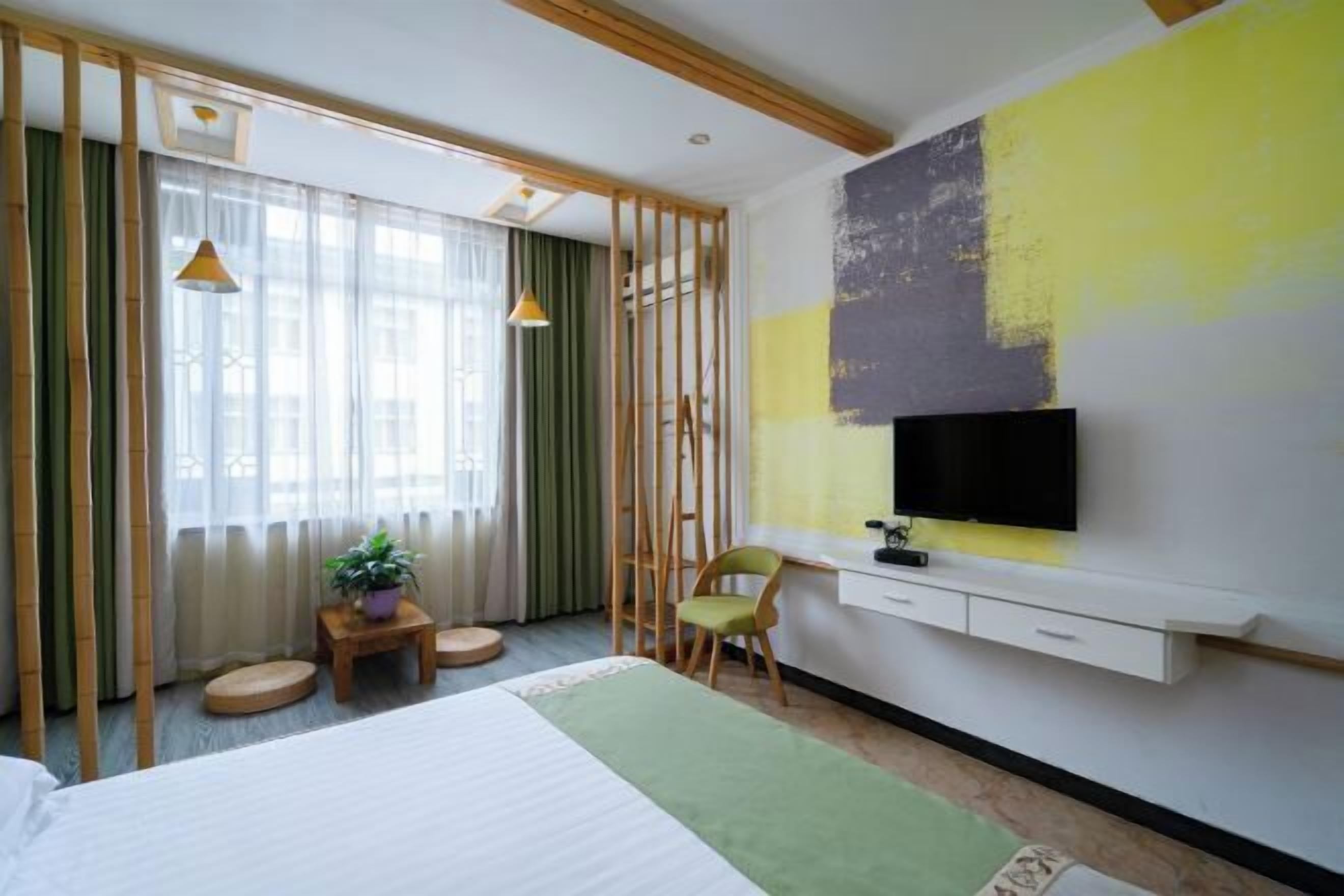 Quarto de hotel com uma cama, uma escrivaninha, uma cadeira, uma televisão e uma janela grande com cortinas.