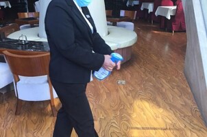 Uma pessoa vestida de terno preto segura uma garrafa de água azul em um saguão moderno de hotel, com piso de madeira e mobiliário branco.