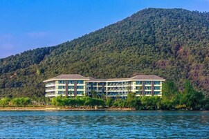 Um hotel de vários andares com design moderno, localizado perto de um corpo de água e com uma colina arborizada ao fundo.