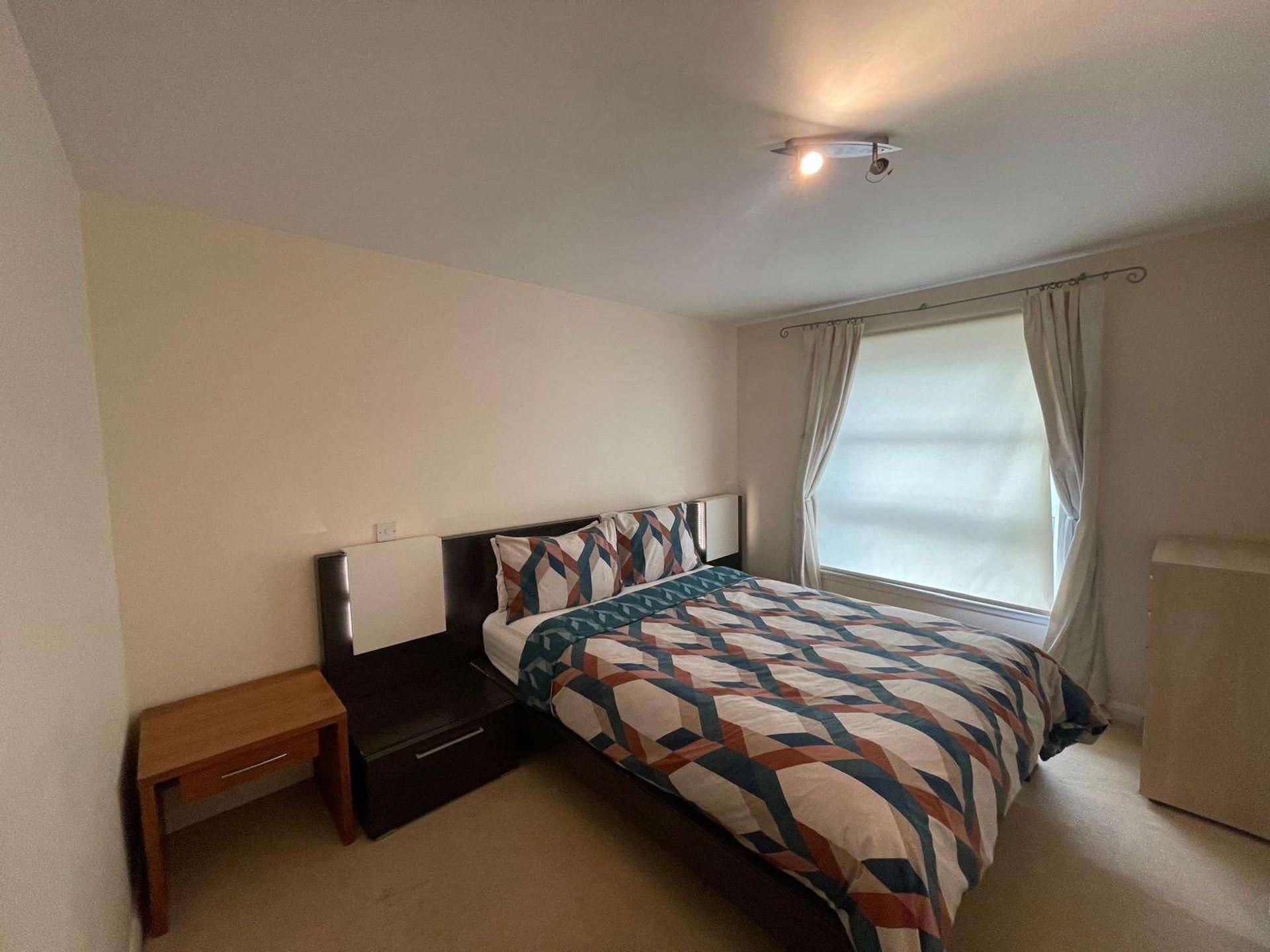 Quarto com cama, mesa de cabeceira, janela com cortinas e luminária no teto.