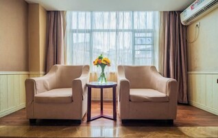 Um quarto com duas poltronas bege, uma mesinha redonda com um vaso de flores e uma janela grande com cortinas.