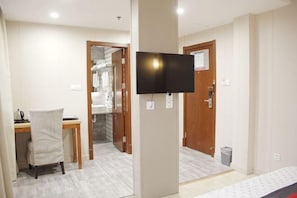 Quarto de hotel com televisão na parede, uma mesa com cadeira e banheiro com chuveiro, além de uma porta.