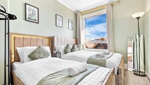 Quarto de hotel com duas camas, uma janela grande, um abajur e uma pequena mesa com uma planta.