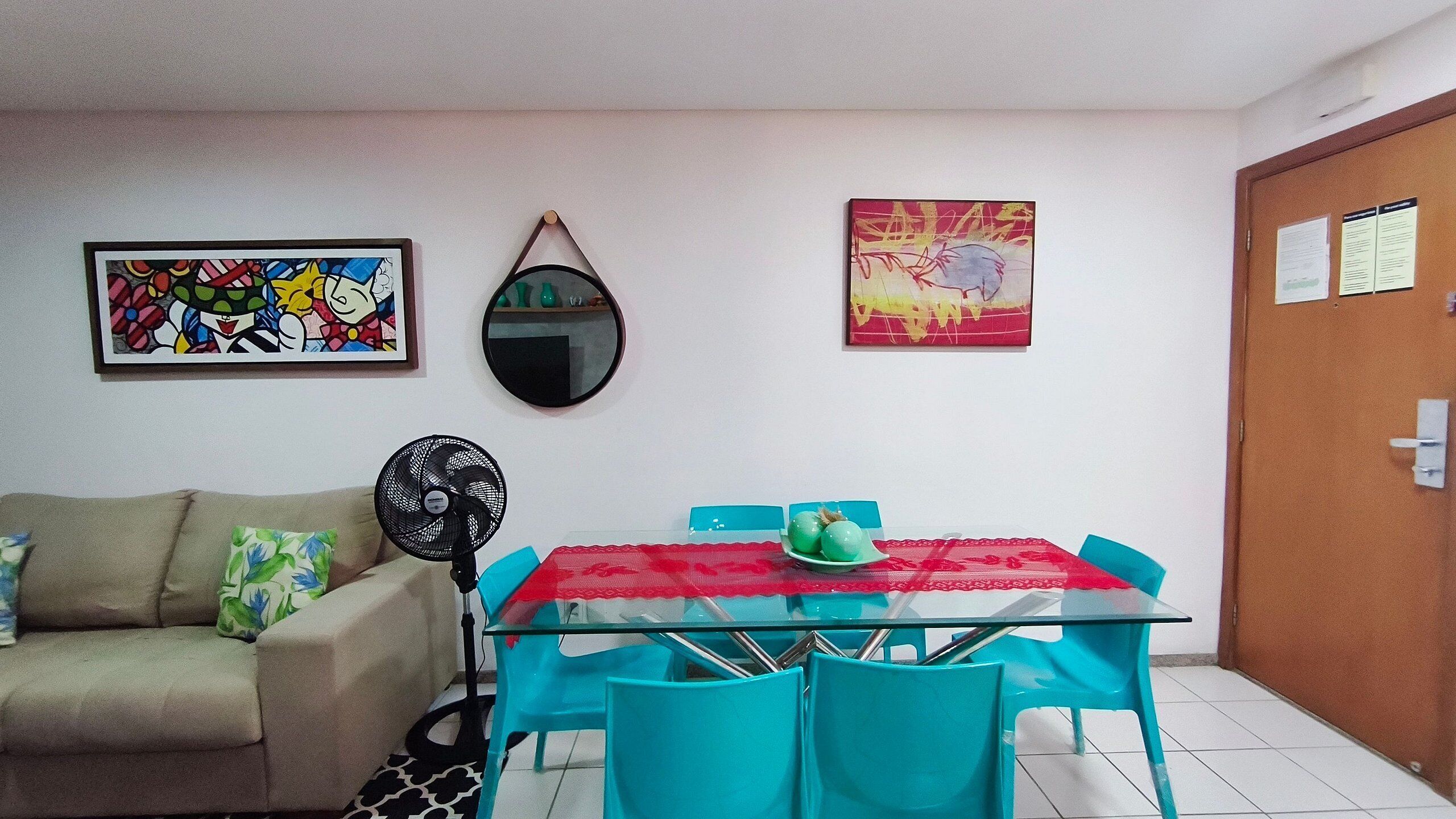 Uma sala de estar com um sofá, uma mesa de vidro com cadeiras, um ventilador e duas obras de arte na parede.