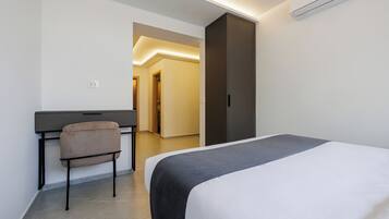 Quarto de hotel com uma cama, uma escrivaninha e uma cadeira. Há também um guarda-roupa e um aparelho de ar condicionado na parede.